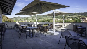 Um pátio externo com mesas, cadeiras, guarda-sóis e vista para o campo.