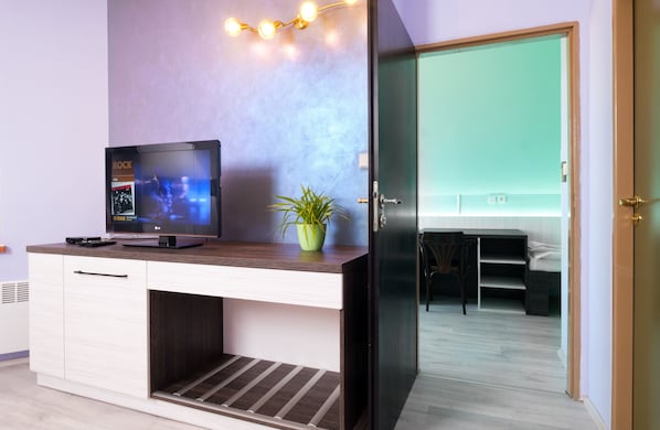 Quarto moderno com televisão de tela plana sobre uma escrivaninha de madeira, uma planta em vaso e uma porta de vidro deslizante que dá para um quarto com cama e escrivaninha.