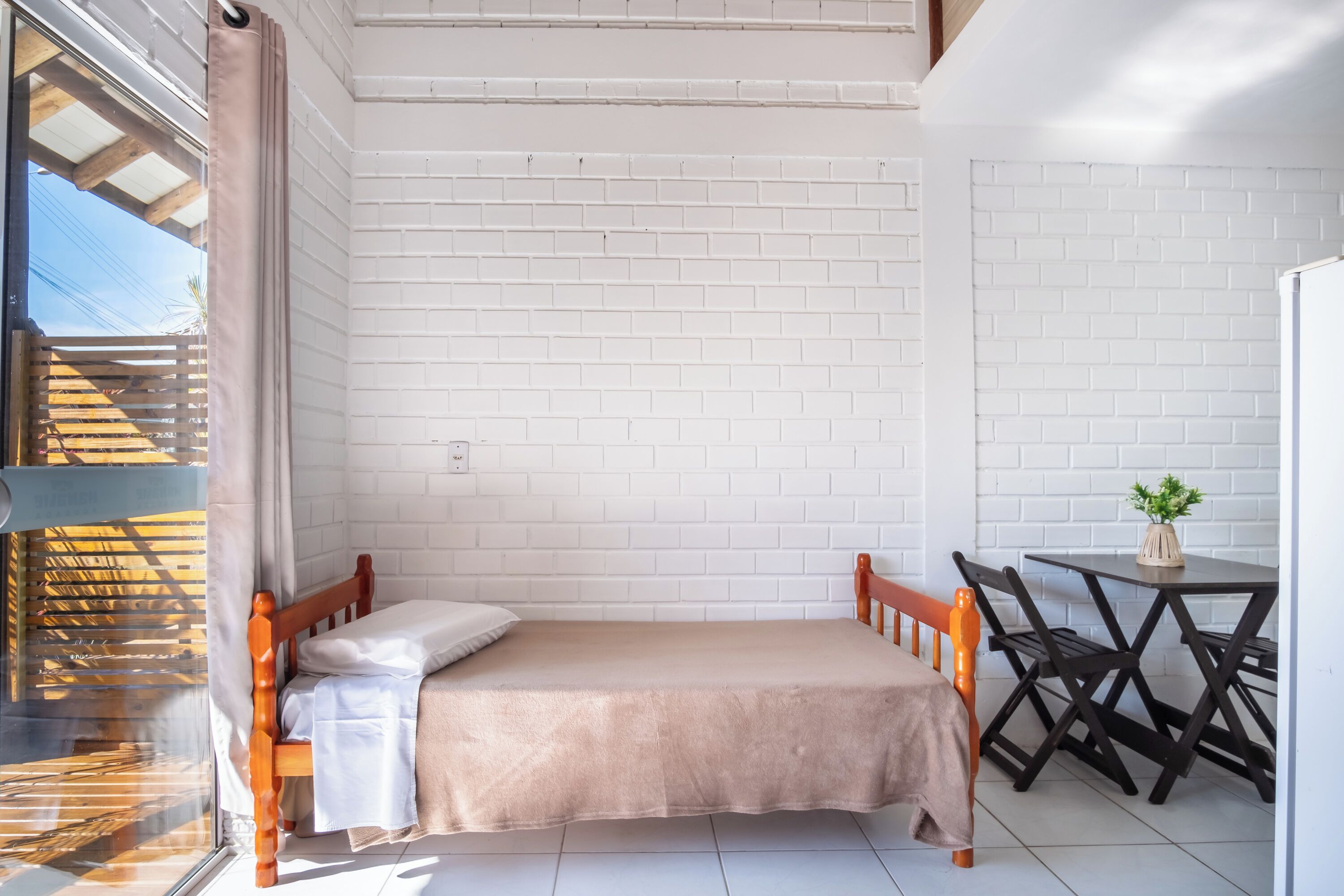 Quarto pequeno com uma cama de solteiro, cabeceira de madeira, uma mesinha com cadeiras e um vaso com planta.
