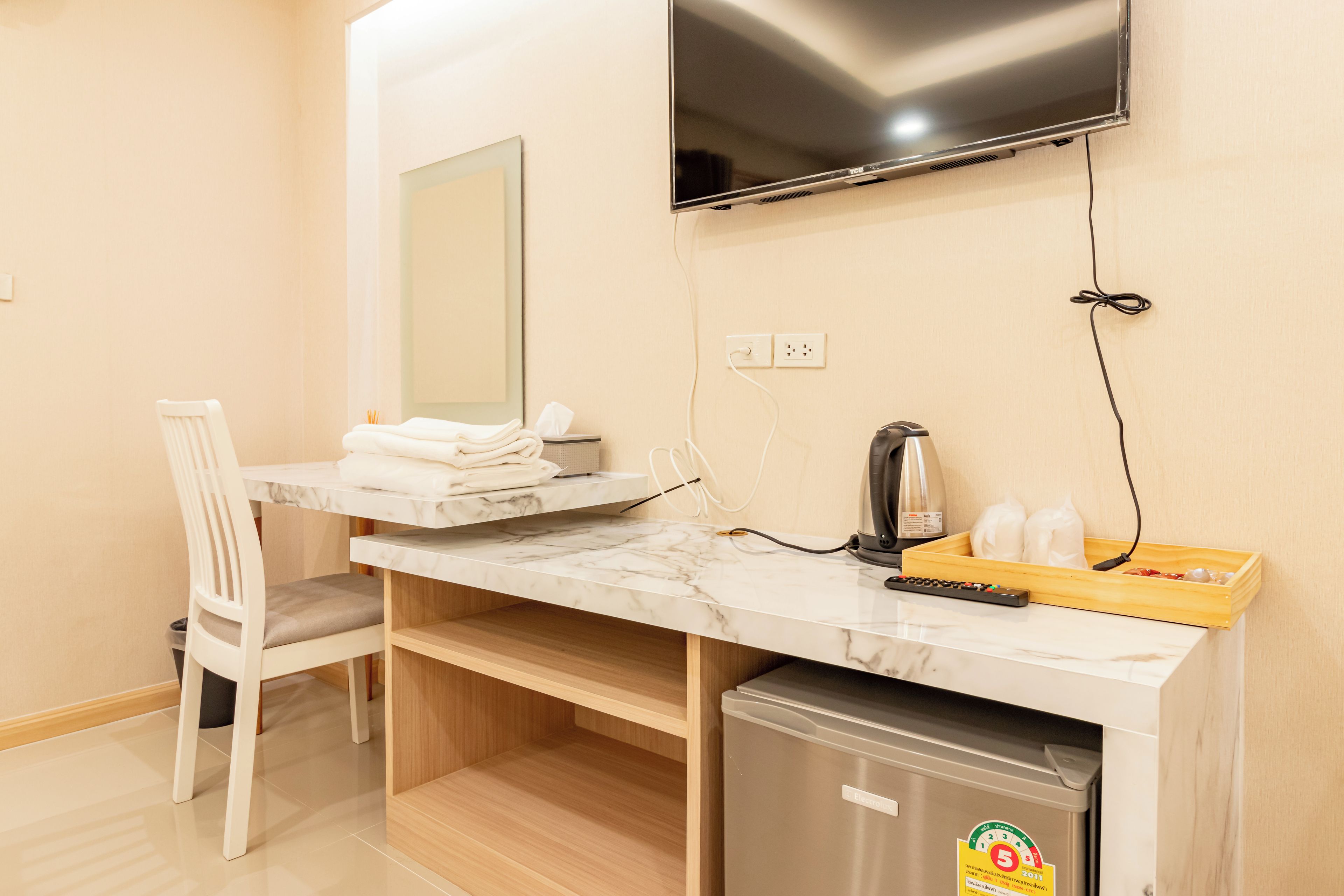 Quarto de hotel com bancada de mármore, televisão montada na parede, pequena cozinha compacta com geladeira e uma cadeira de madeira.
