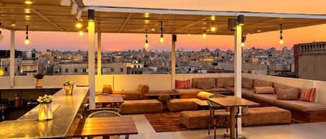 Terraço no telhado com área de bar, assentos e vista da cidade ao pôr do sol.