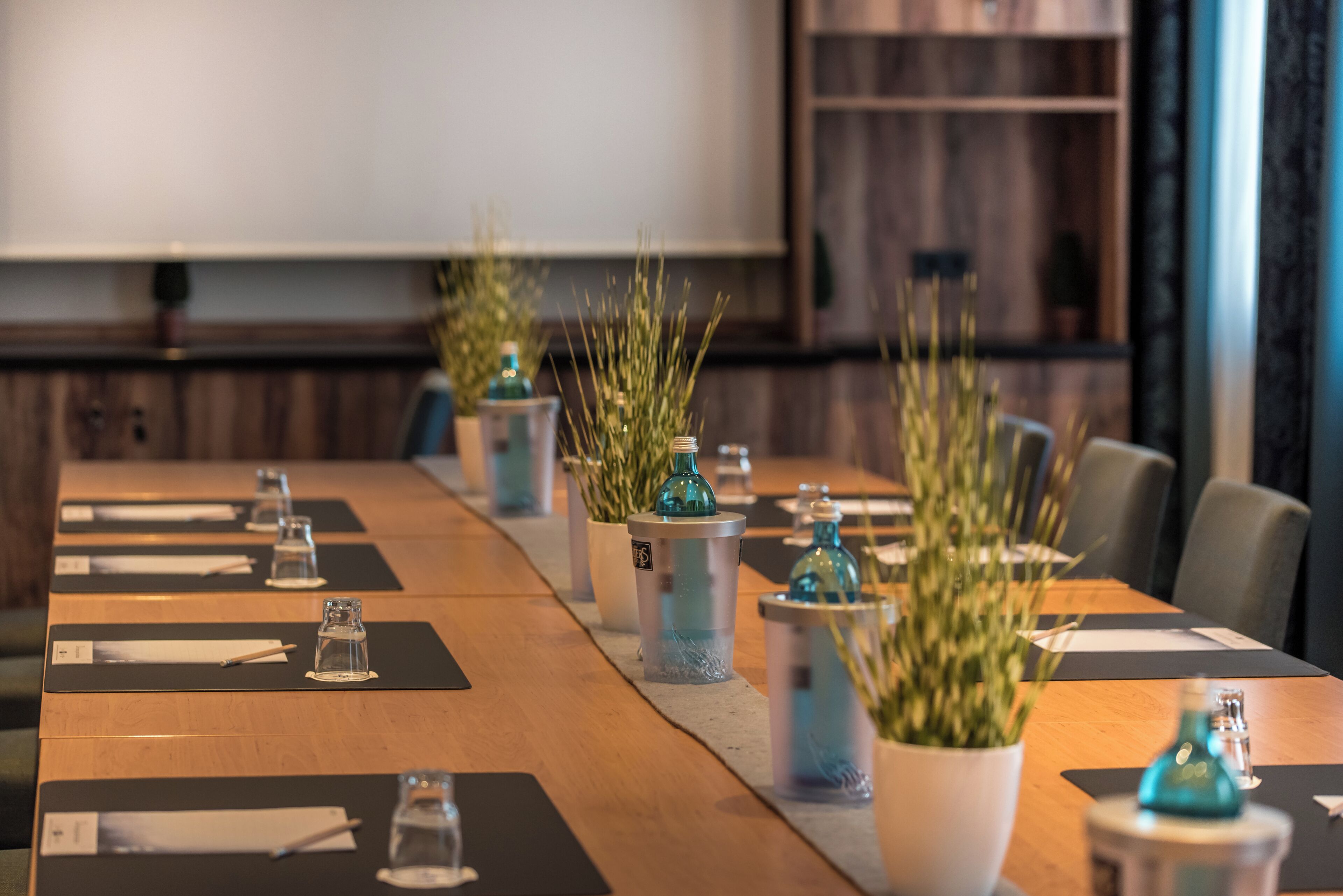 Uma sala de conferências com uma mesa comprida, plantas em vasos e uma tela de projetor ao fundo.