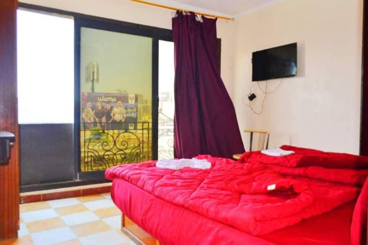 Quarto com colcha vermelha, televisão na parede e vista para uma varanda com um pôster.