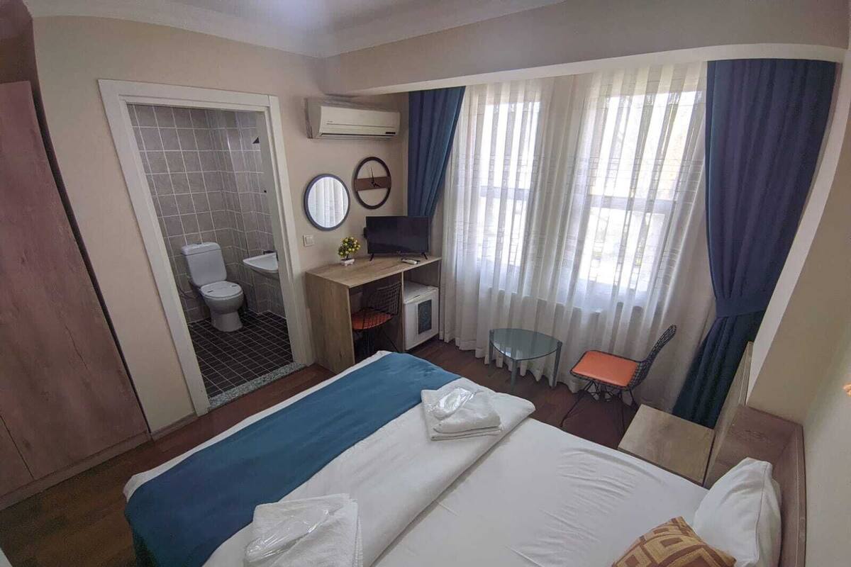 Quarto de hotel com cama, escrivaninha, cadeira, mesa pequena, espelho e janela com cortinas.