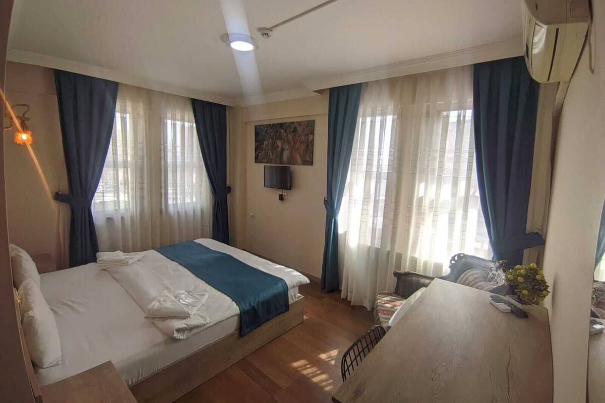 Quarto de hotel com cama, escrivaninha, cadeira, televisão e um quadro na parede.