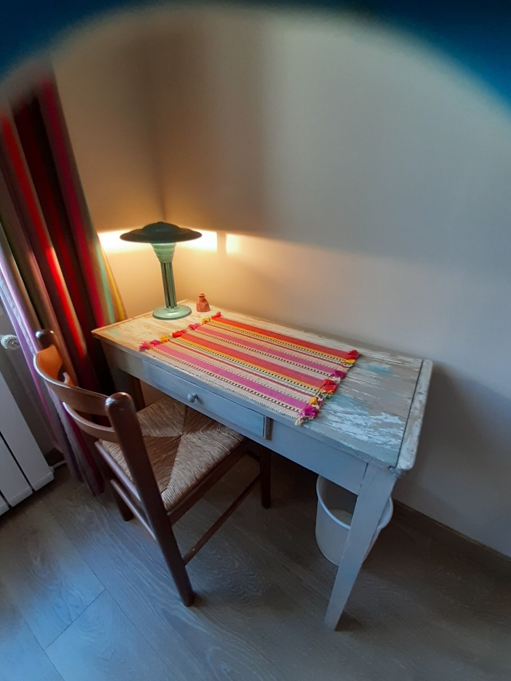 Uma pequena mesa de madeira com um jogo de americano tecido colorido, uma lâmpada e uma cadeira.