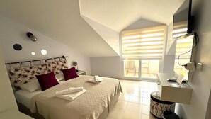Quarto com cama, televisão de tela plana, janela com persianas e pequena mesa com abajur.