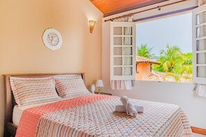 Cama bem arrumada com colcha rosa e branca, toalha branca sobre a cama, e janela com cortinas abertas para uma vista de palmeiras e um prédio.