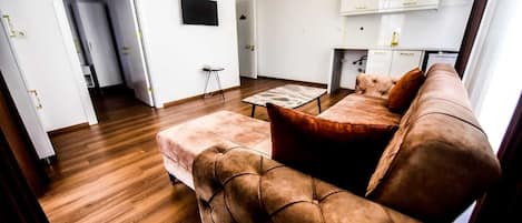 Uma sala de estar moderna com um sofá e um banquinho marrons, piso de madeira, uma mesinha e uma cozinha ao fundo.