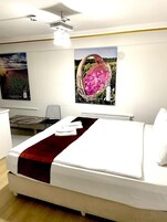 Quarto de hotel com uma cama, uma escrivaninha e uma cadeira. Há dois cartazes na parede, um deles com um cesto de flores.