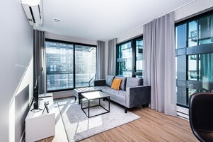 Sala de estar moderna com um sofá cinzento, uma mesa de centro e uma TV em um móvel. Janelas amplas proporcionam vista da cidade.