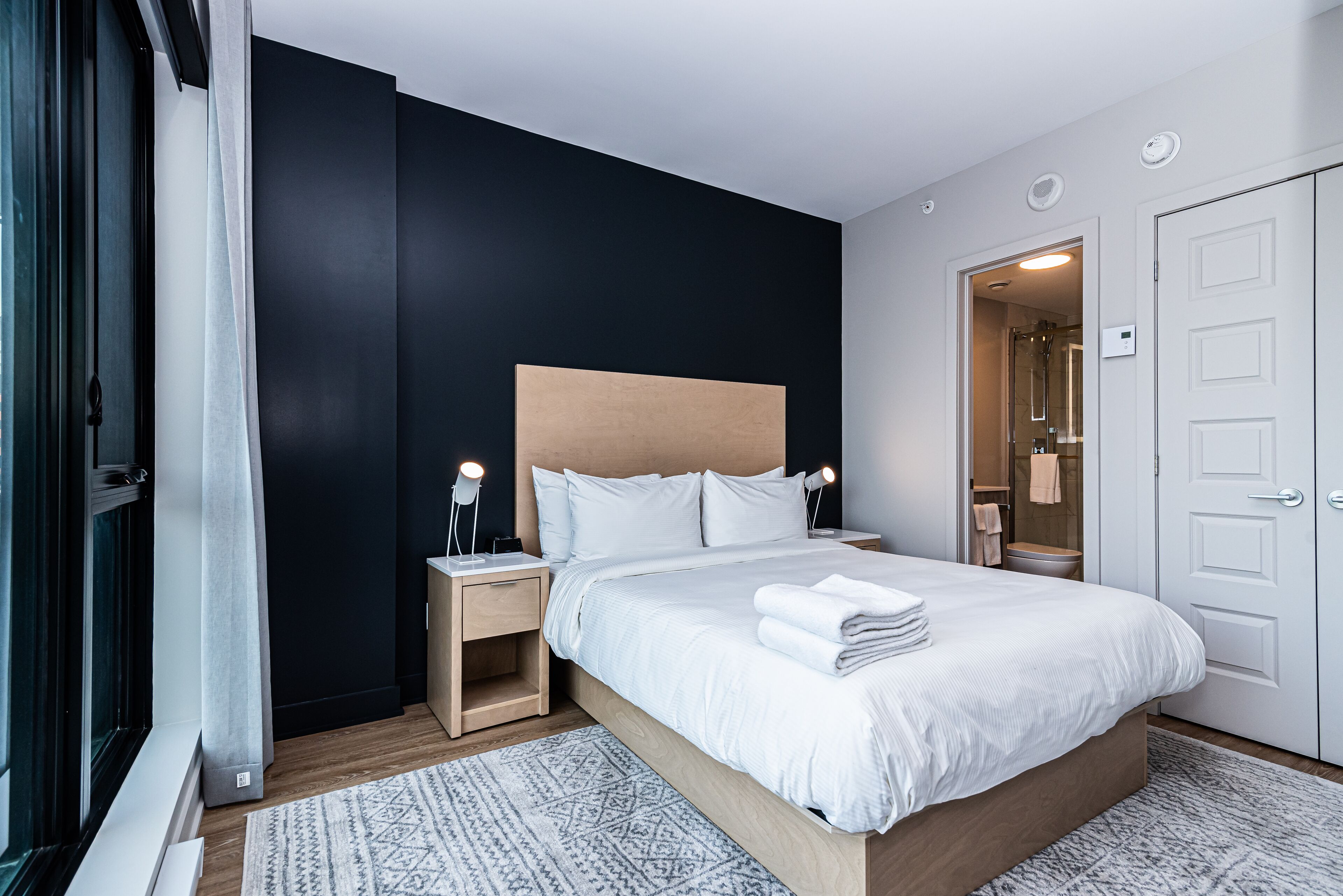 Quarto de hotel moderno com uma cama grande, mesas de cabeceira com abajures, um guarda-roupa e um banheiro visível por uma porta aberta.