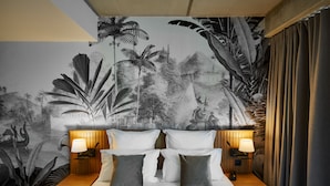 Quarto de hotel com uma cama grande, duas mesas de cabeceira com luminárias e um mural com paisagem tropical na parede.
