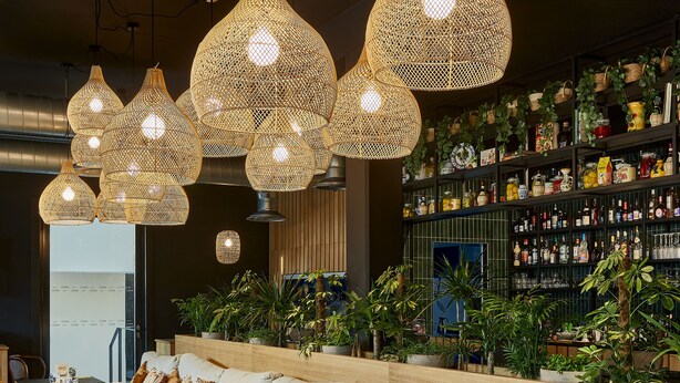 Um restaurante com uma longa mesa de refeições comunitária, luminárias pendentes de vime e diversas plantas.