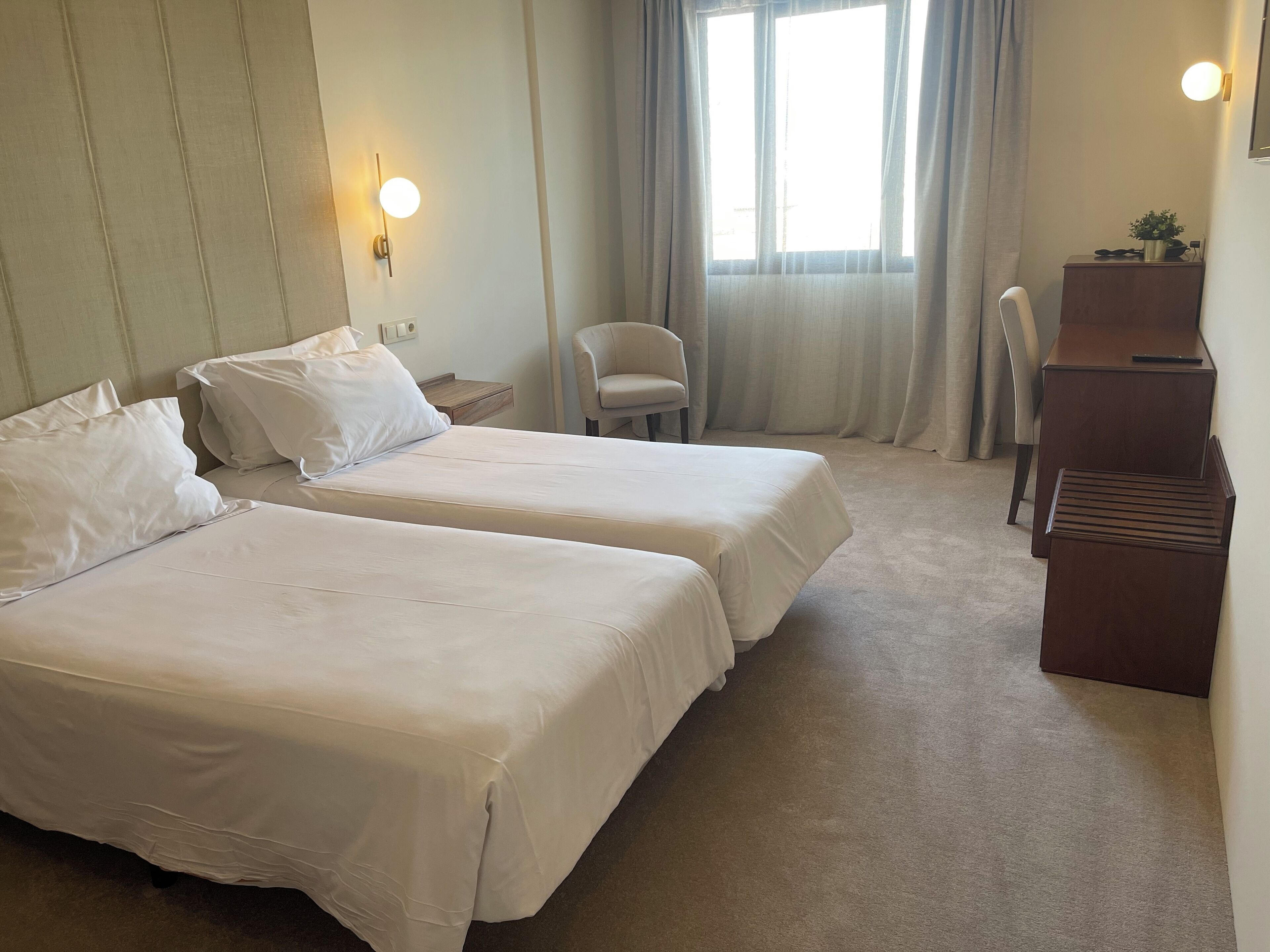 Quarto de hotel com duas camas, uma escrivaninha, uma cadeira e uma janela com cortinas.