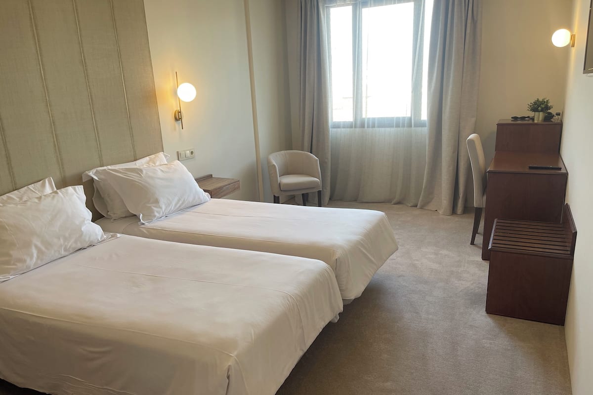 Quarto de hotel com duas camas, uma escrivaninha, uma cadeira e uma janela com cortinas.