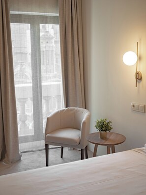 Quarto de hotel com cama, uma cadeira, uma mesinha com uma planta e uma luminária na parede.