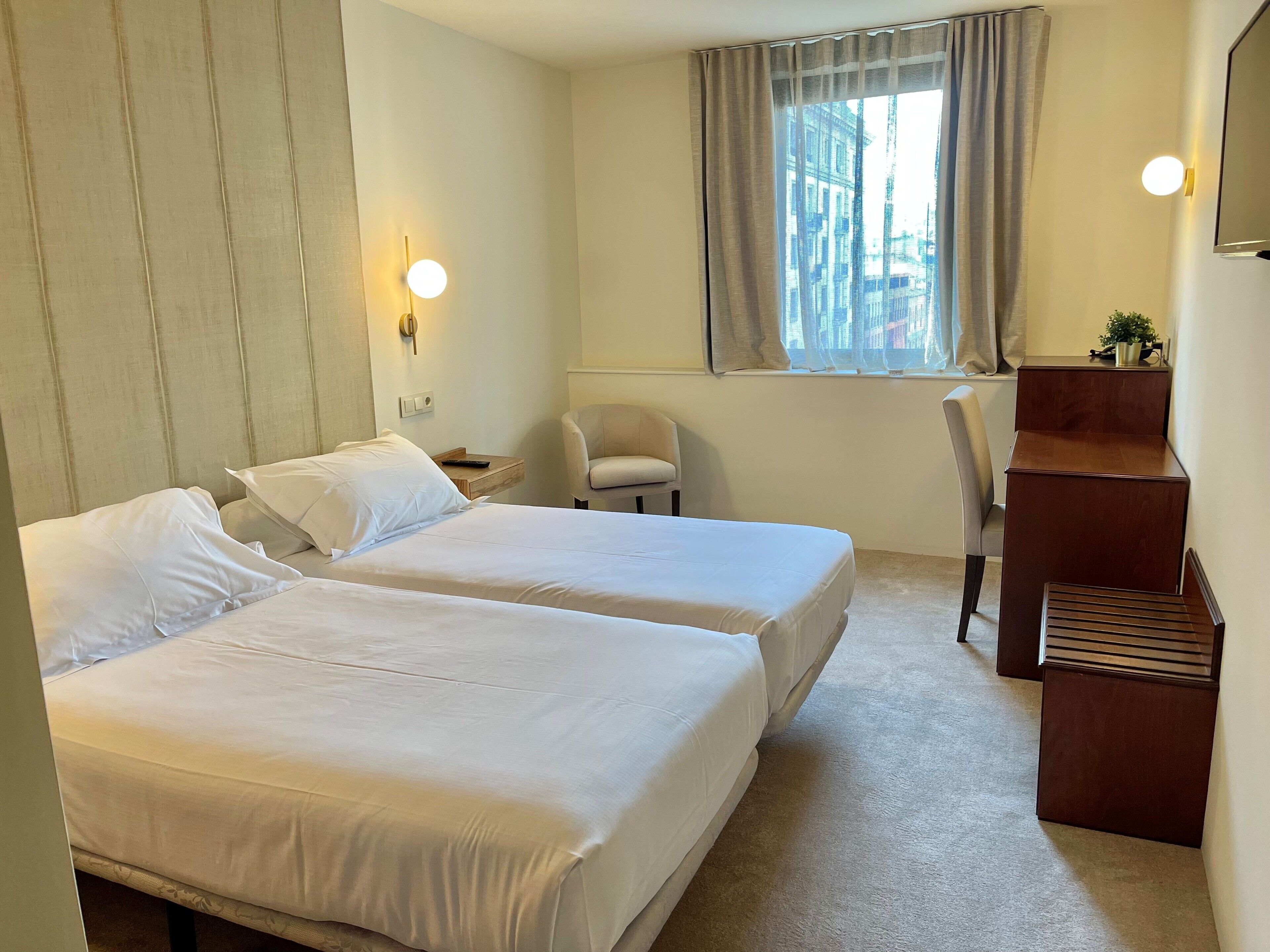 Quarto de hotel com duas camas, uma escrivaninha, uma cadeira, uma janela com cortinas e televisão na parede.