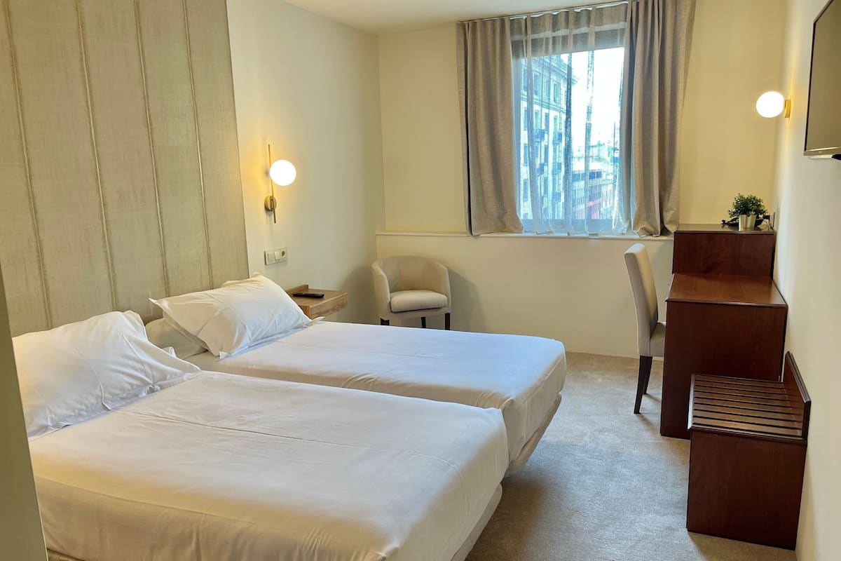 Quarto de hotel com duas camas, uma escrivaninha, uma cadeira, uma janela com cortinas e televisão na parede.
