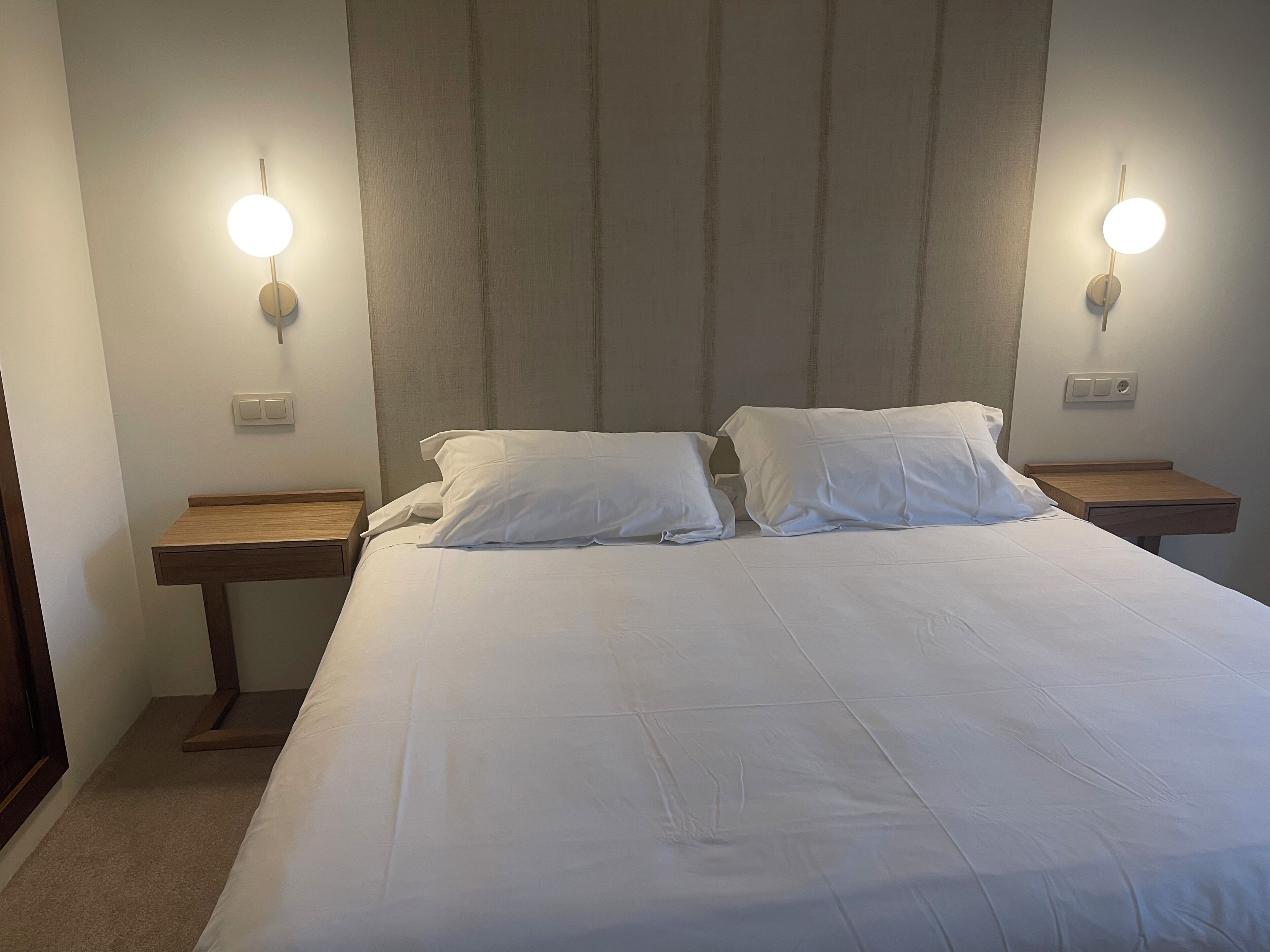 Uma cama bem arrumada com lençóis brancos, duas mesas de cabeceira com luminárias e uma parede lisa com um interruptor de luz.