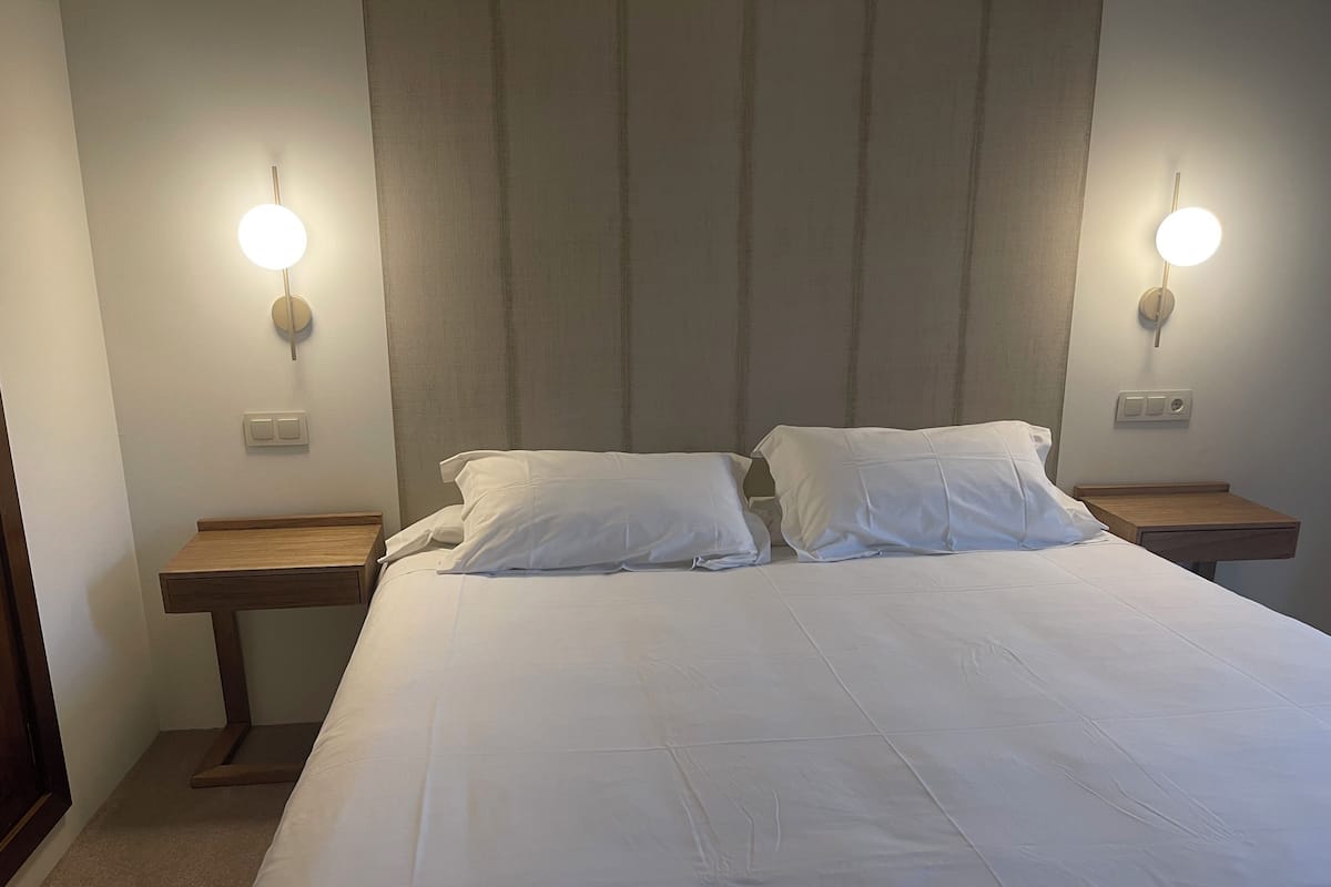 Cama bem arrumada com lençóis brancos, duas mesas de cabeceira com abajures e cabeceira em tom neutro.