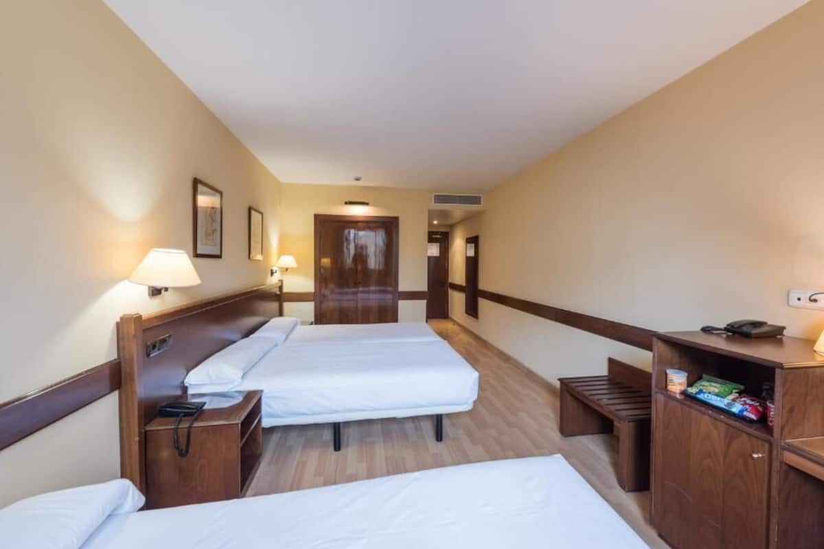 Quarto de hotel com duas camas de solteiro, uma escrivaninha de madeira, uma cadeira, uma pequena prateleira com artigos de higiene pessoal e uma porta que dá para outro cômodo.