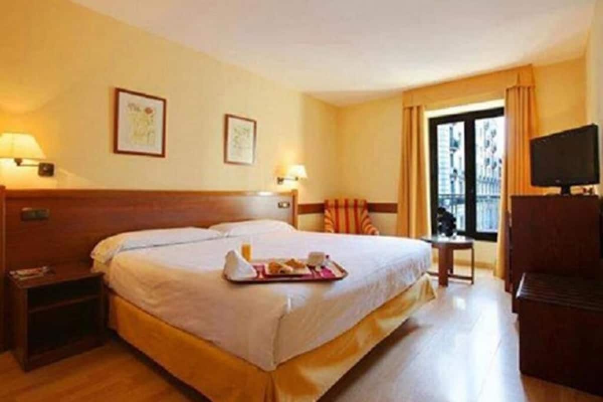 Quarto de hotel com uma cama grande, mesas de cabeceira, televisão e janela com cortinas.