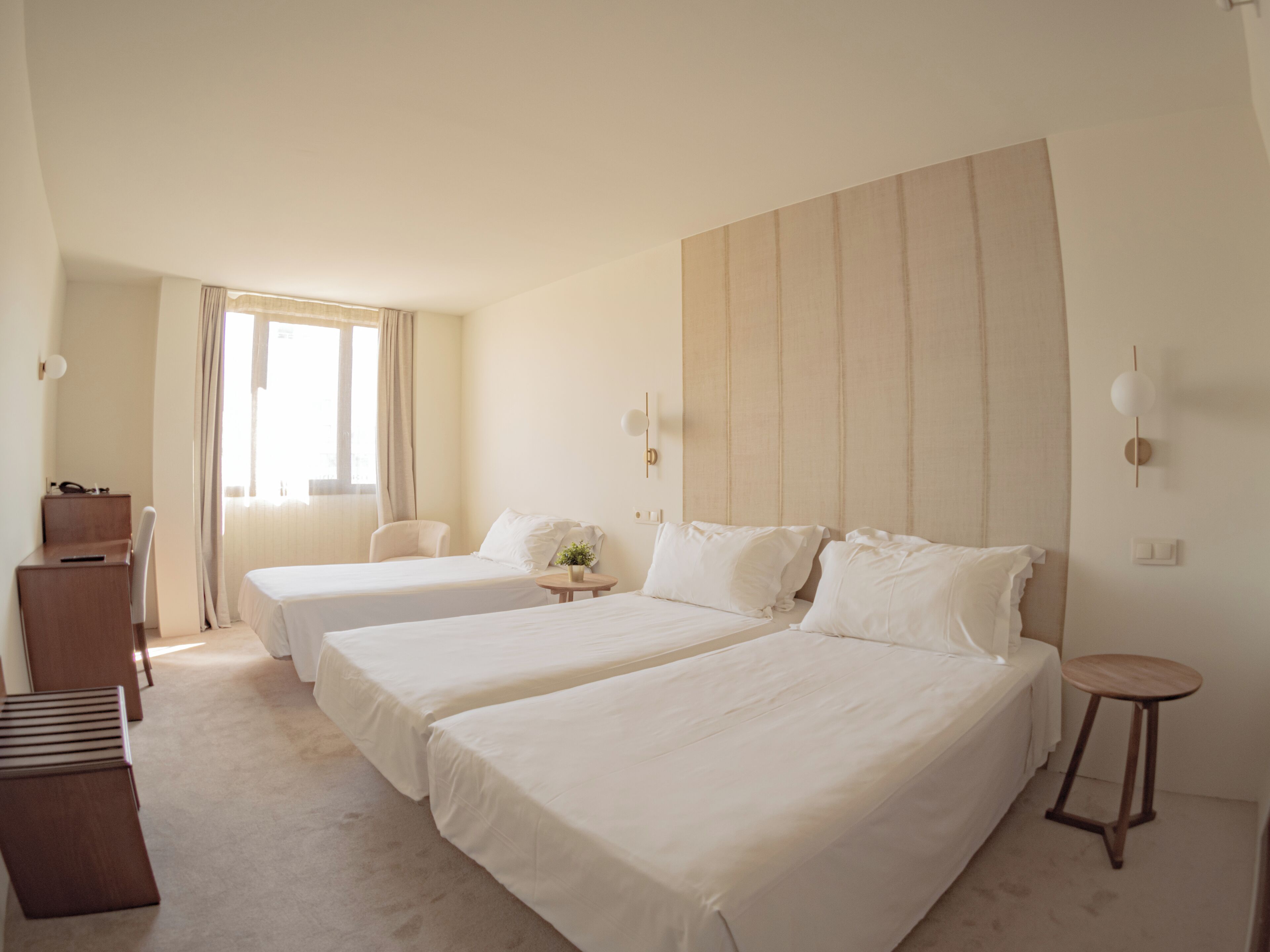 Quarto de hotel com duas camas, uma escrivaninha, uma cadeira e uma pequena mesa com uma planta.