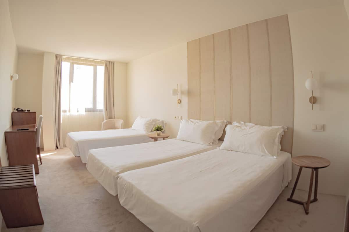 Quarto de hotel com duas camas, uma escrivaninha, uma cadeira, uma mesinha e uma janela com cortinas.