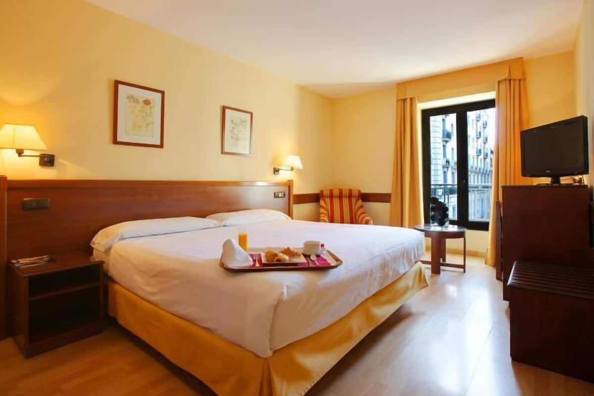 Quarto de hotel com uma cama grande, luminárias de cabeceira, televisão e uma janela com cortinas.
