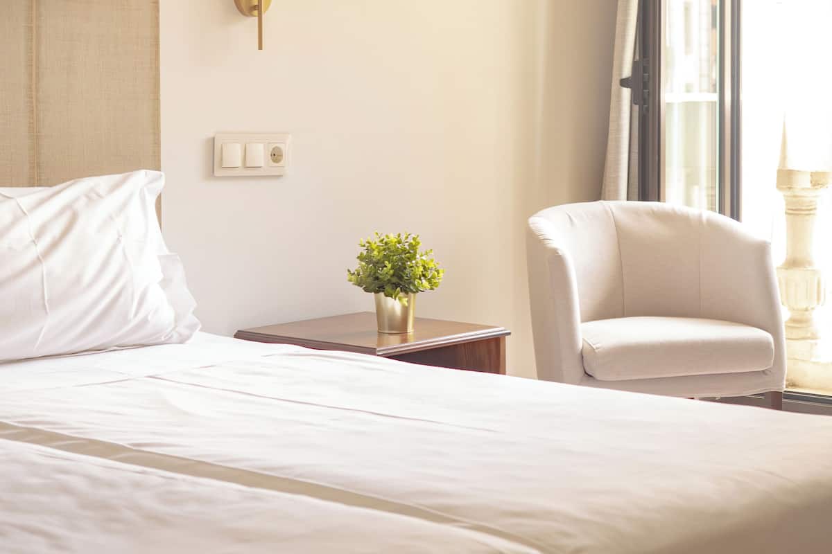 Quarto de hotel com uma cama, uma cadeira, luminária fixada na parede e janela com cortinas.