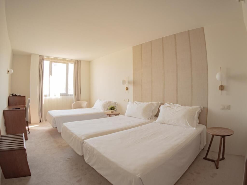 Quarto de hotel com duas camas, uma escrivaninha, uma cadeira, uma mesinha e uma janela com cortinas.