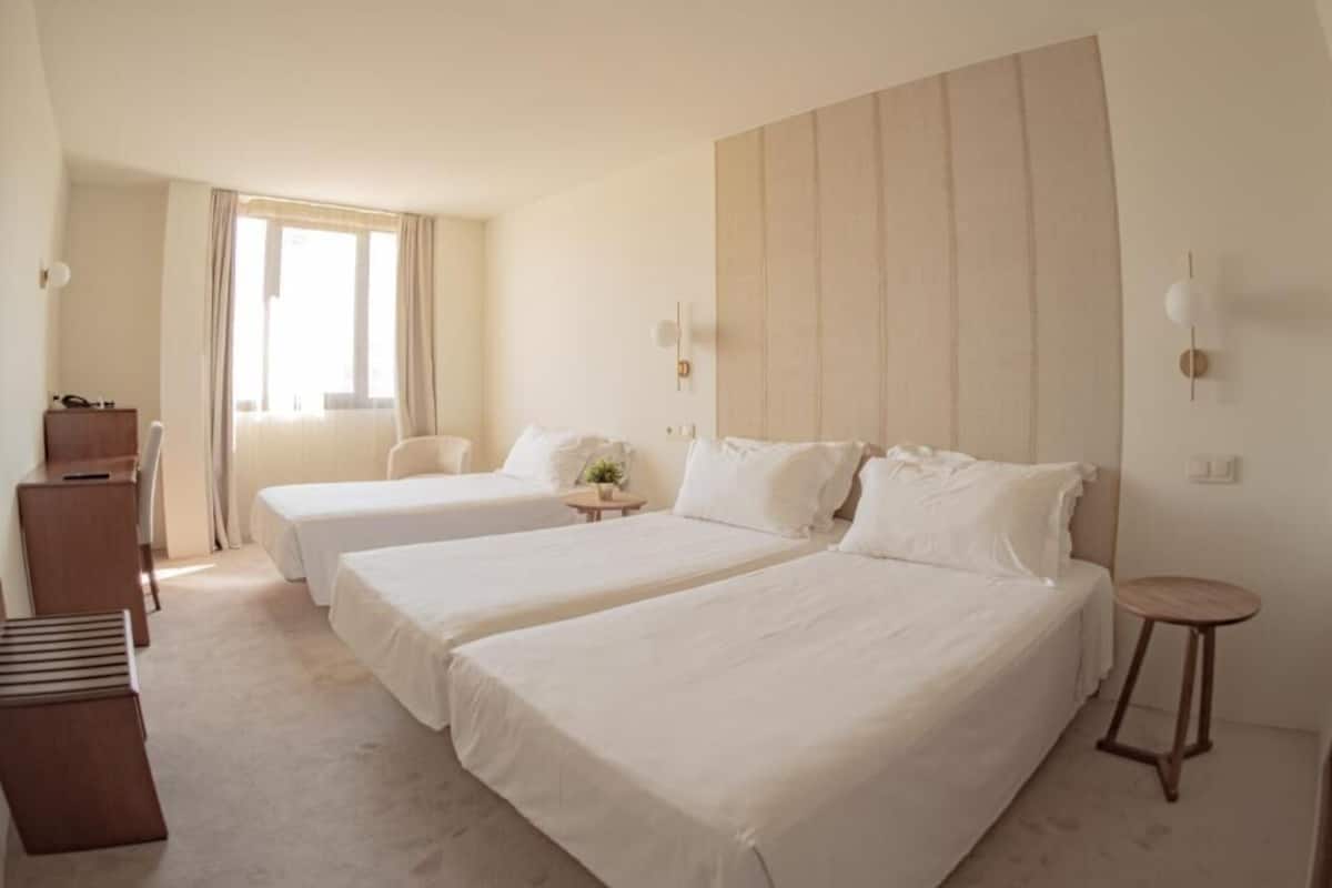 Quarto de hotel com duas camas, uma escrivaninha, uma cadeira, uma mesinha e uma janela com cortinas.