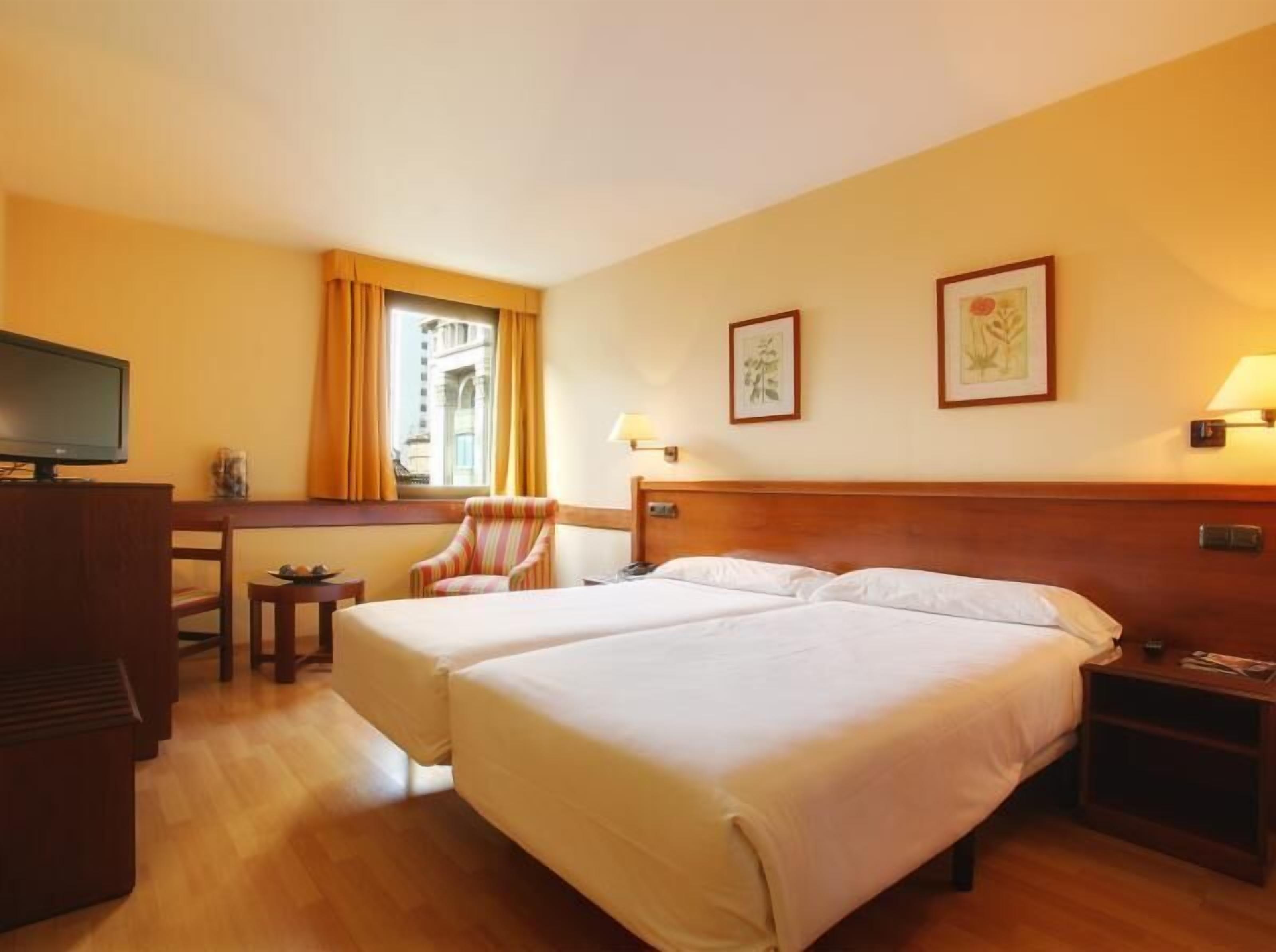 Quarto de hotel com duas camas, uma escrivaninha com cadeira, televisão e obras de arte nas paredes.