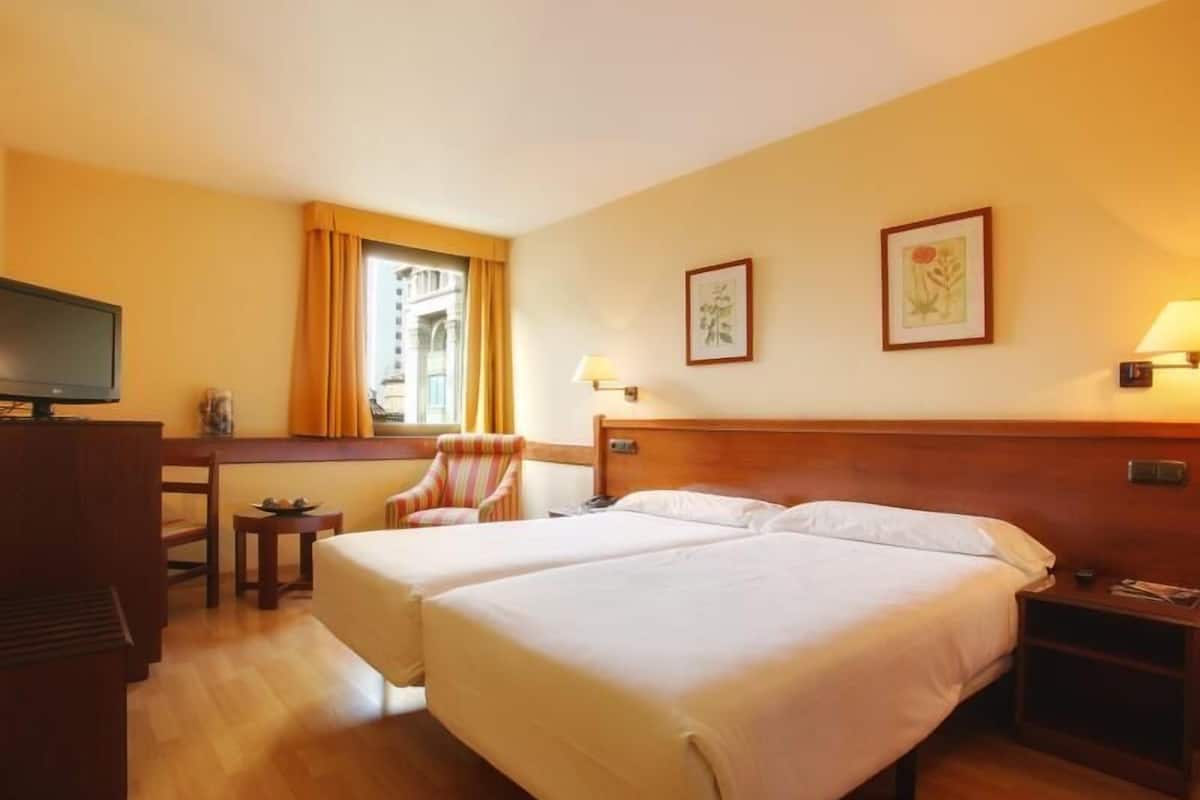 Quarto de hotel com duas camas, uma escrivaninha com cadeira, televisão e obras de arte nas paredes.