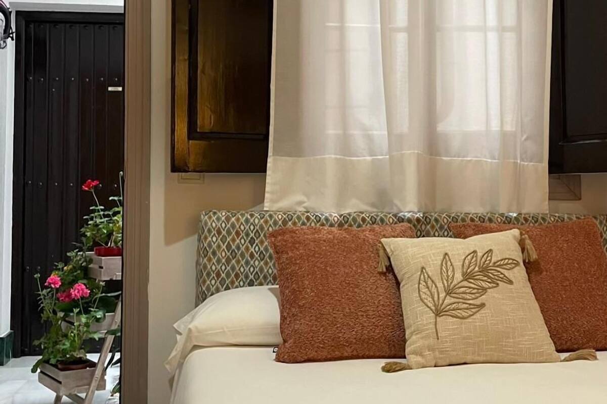 Um quarto com cama, janela com cortinas leves, porta e planta em vaso com flores.