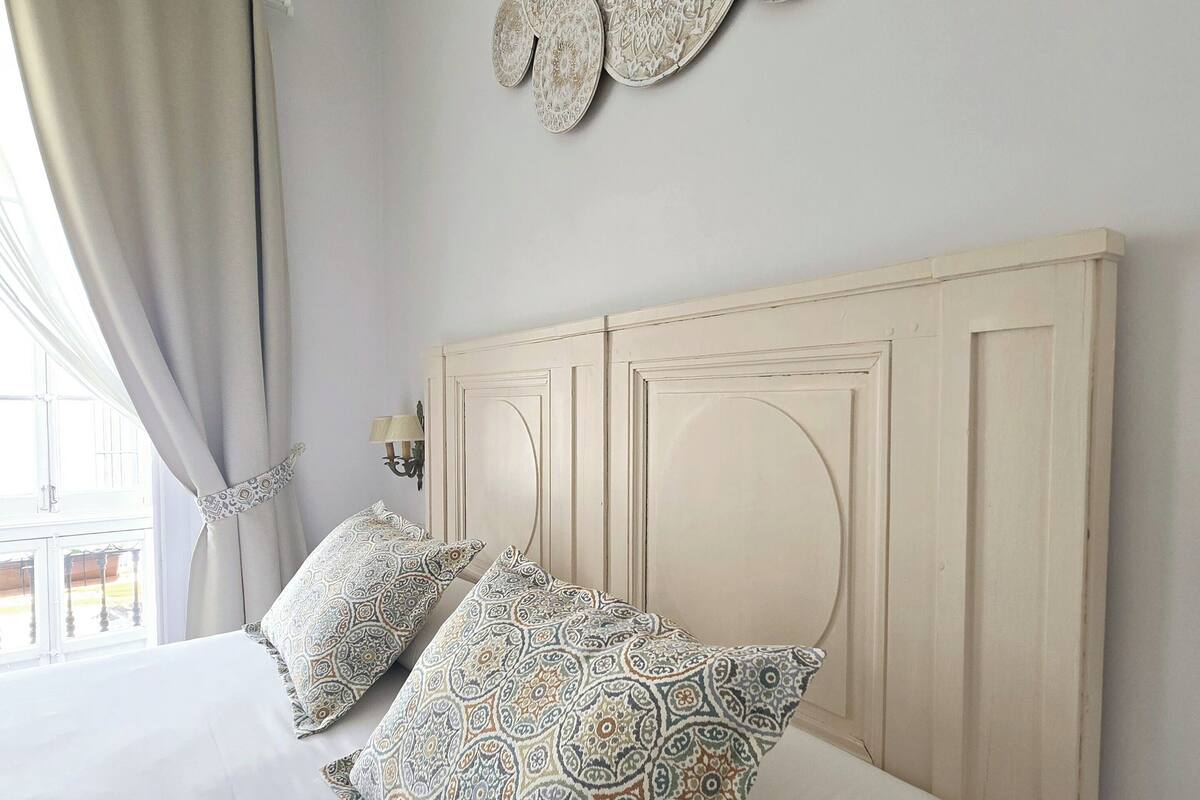 Quarto com cama grande, almofadas estampadas, um objeto decorativo na parede e uma janela com cortinas.
