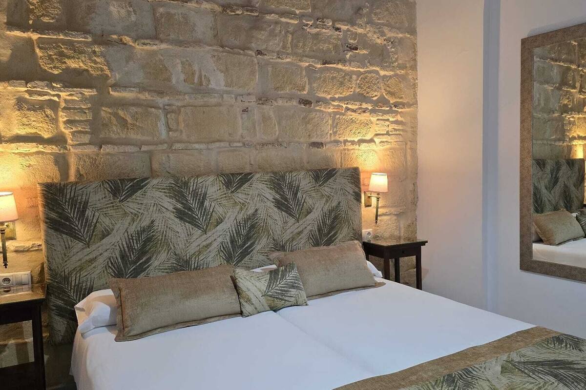 Quarto com parede de pedra, cama com cabeceira estampada com folhas, duas luminárias de cabeceira e um espelho na parede.