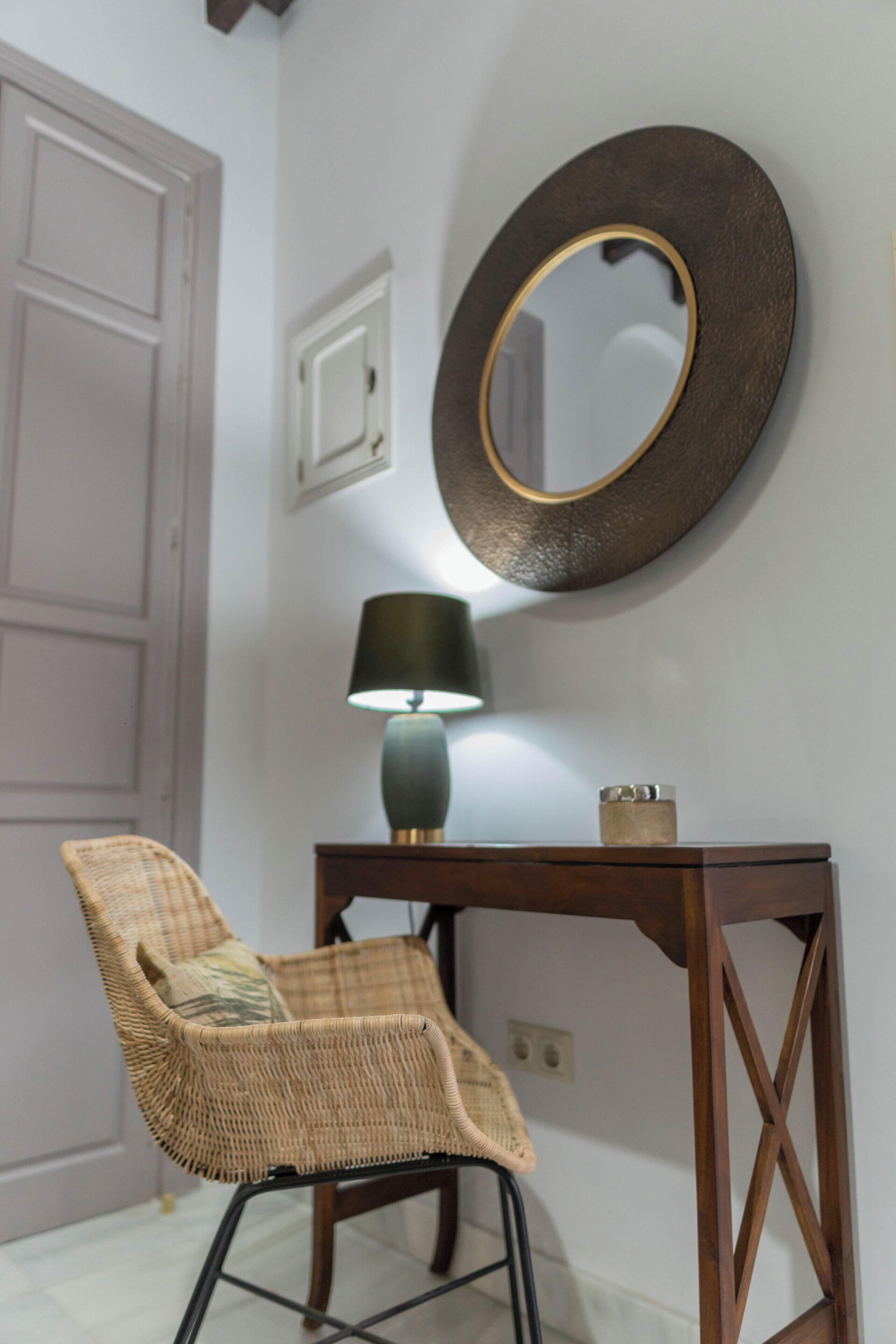 Um quarto com uma cadeira de vime, uma mesa com abajur e um espelho fixado na parede.