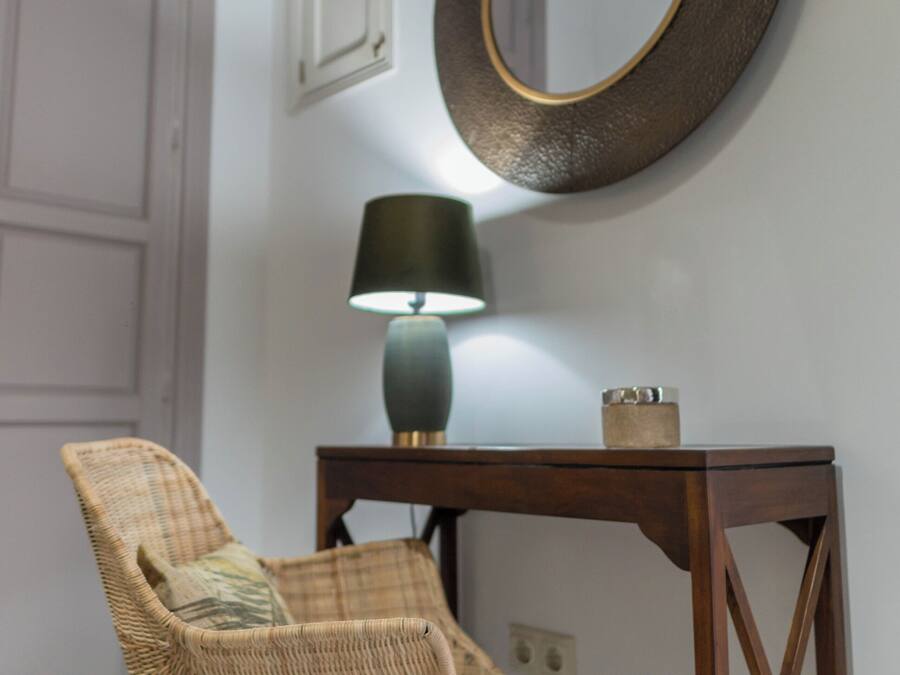 Um quarto com uma cadeira de vime, uma mesa com abajur e um espelho fixado na parede.