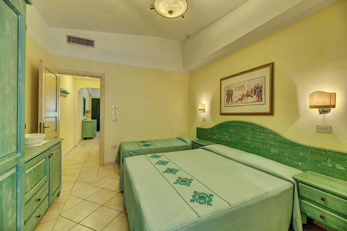 Quarto com duas camas, um penteou verde, um quadro na parede e porta para outro cômodo.