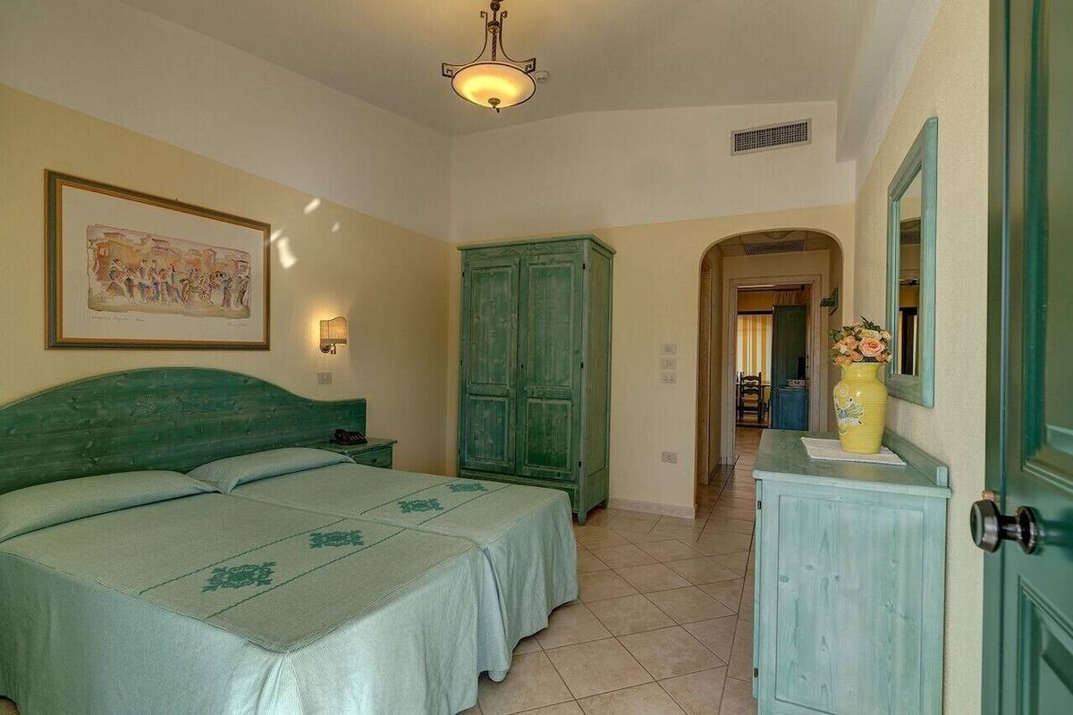 Quarto com cama, guarda-roupa verde, mesa de cabeceira, quadro na parede e porta para outro cômodo.