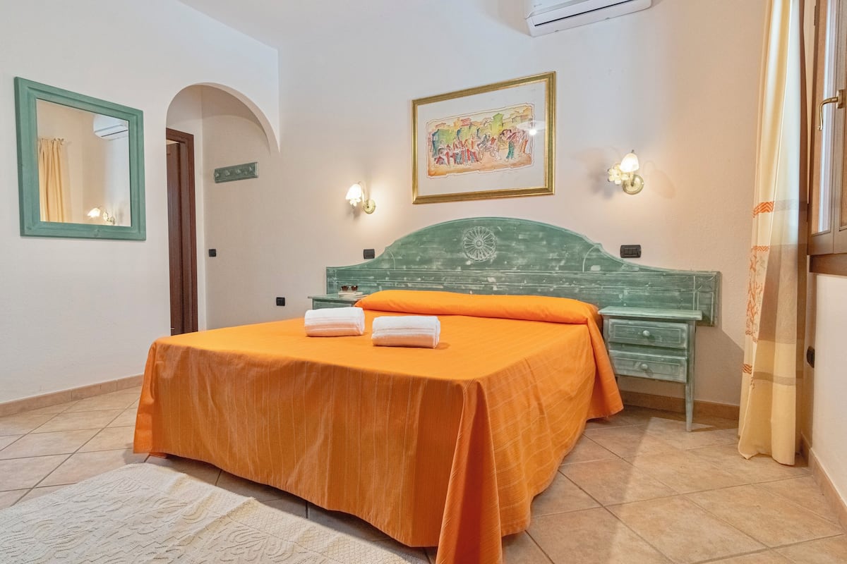 Quarto com cabeceira verde, colcha laranja, duas toalhas brancas, mesa de cabeceira, gaveta, luminária na parede, quadro emoldurado e espelho com moldura verde.