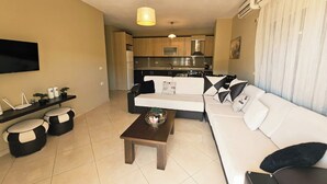 Uma sala de estar moderna com um sofá, mesa de centro e televisão. Uma cozinha fica ao fundo.