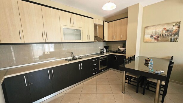 Cozinha moderna com armários preto e bege, pia de aço inoxidável e área de jantar com mesa de vidro e cadeiras.
