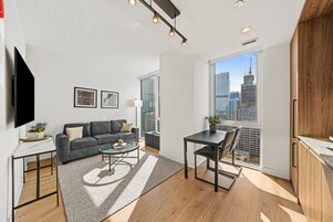 Uma sala de estar moderna com sofá, mesa de centro e mesa de jantar. Há uma TV fixada na parede, uma janela com vista para a cidade e um armário de madeira.