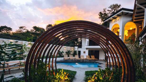 Uma área de piscina com uma escultura metálica única formando um arco sobre a passagem, cercada por vegetação exuberante e um edifício com o nome "ONDA" visível.