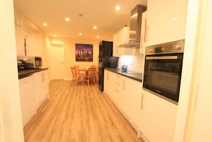 Uma cozinha compacta com armários brancos, uma geladeira preta, micro-ondas e forno. Há uma mesa de jantar com cadeiras e uma pintura de paisagem urbana na parede.