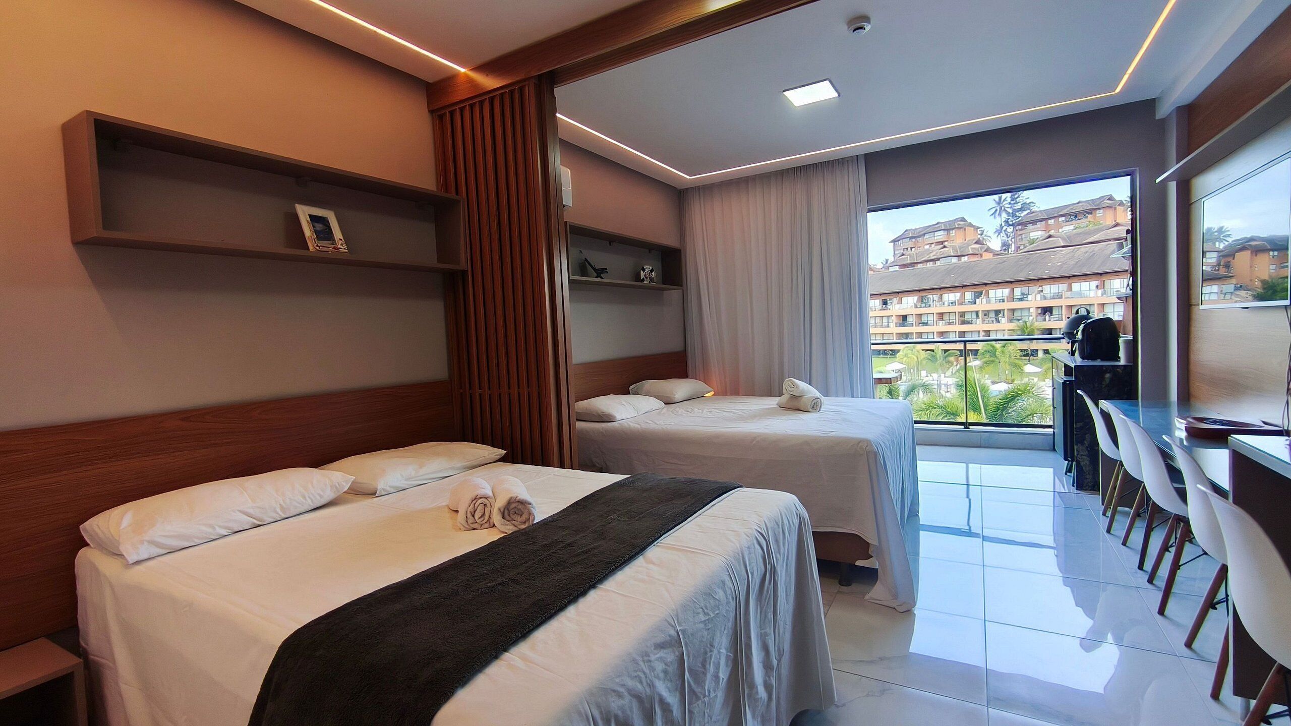 Quarto de hotel com duas camas, uma escrivaninha, uma cadeira, uma janela com vista para edifícios e iluminação embutida no teto.