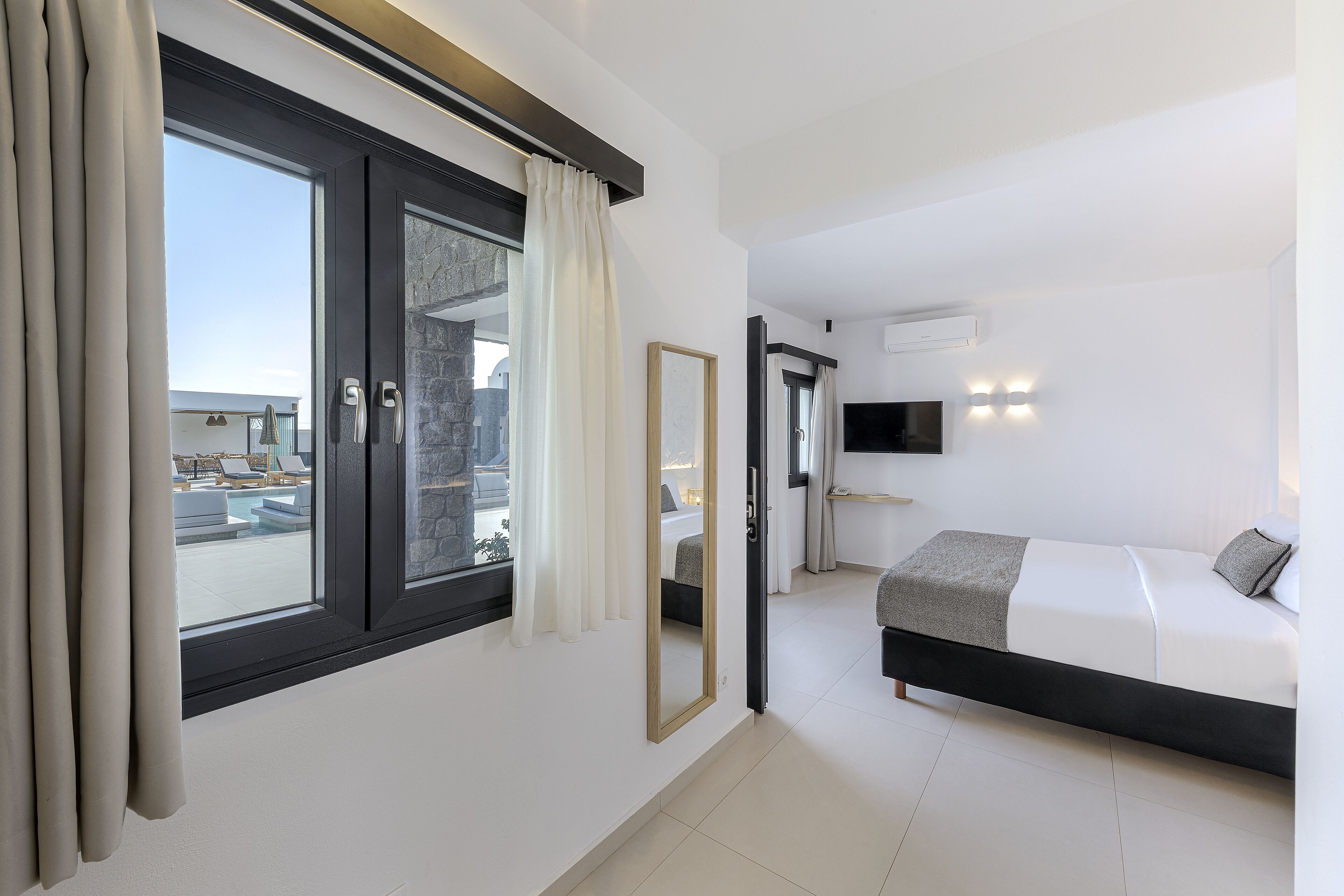 Quarto de hotel moderno com uma janela grande, uma cama, uma mesa de cabeceira e uma televisão fixada na parede.